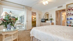 Quarto de hotel com uma cama grande, uma mesa de vidro com um vaso de flores, televisão e estante de livros.
