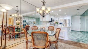 Uma sala de jantar com uma mesa de vidro, rodeada por cadeiras de vime, um candelabro e um espelho grande que reflete a cozinha.