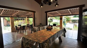 Um espaço de refeições interno com uma mesa comprida coberta por um mantel floral, rodeada por cadeiras. Há duas janelas amplas com vista para um jardim lá fora.