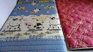 Uma cama com colchão azul decorado com vacas em desenho animado e cobertura acolchoada vermelha.
