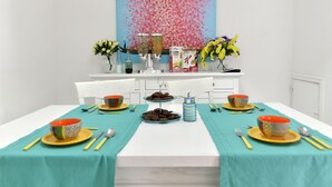 Mesa de jantar posta com tigelas, pratos e talheres coloridos, com uma parede decorativa e um armário branco ao fundo.