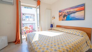 Quarto de hotel com uma cama, varanda com cortinas laranja, quadro com paisagem costeira e vista para edifícios.