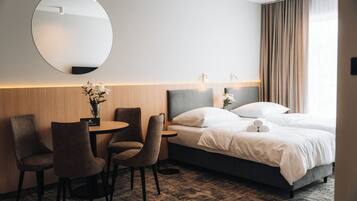 Quarto de hotel com cama, duas cadeiras, uma mesinha, espelho redondo e vaso com flores.