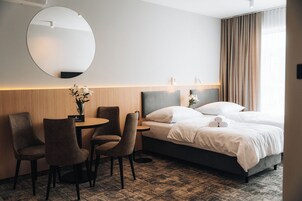 Quarto de hotel com cama, duas cadeiras, uma mesinha, espelho redondo e vaso com flores.