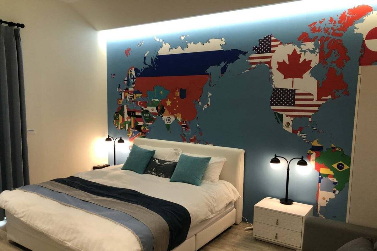 Quarto moderno com um grande mural de mapa mundi, uma cama com manta listrada, duas mesas de cabeceira com luminárias e um sofá cinza.