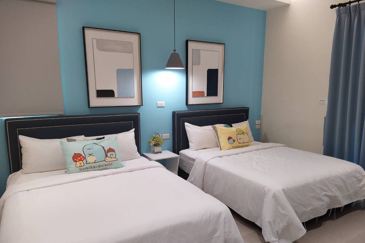 Duas camas de solteiro com roupa de cama branca, uma pequena mesa de cabeceira com abajur e parede com duas molduras.