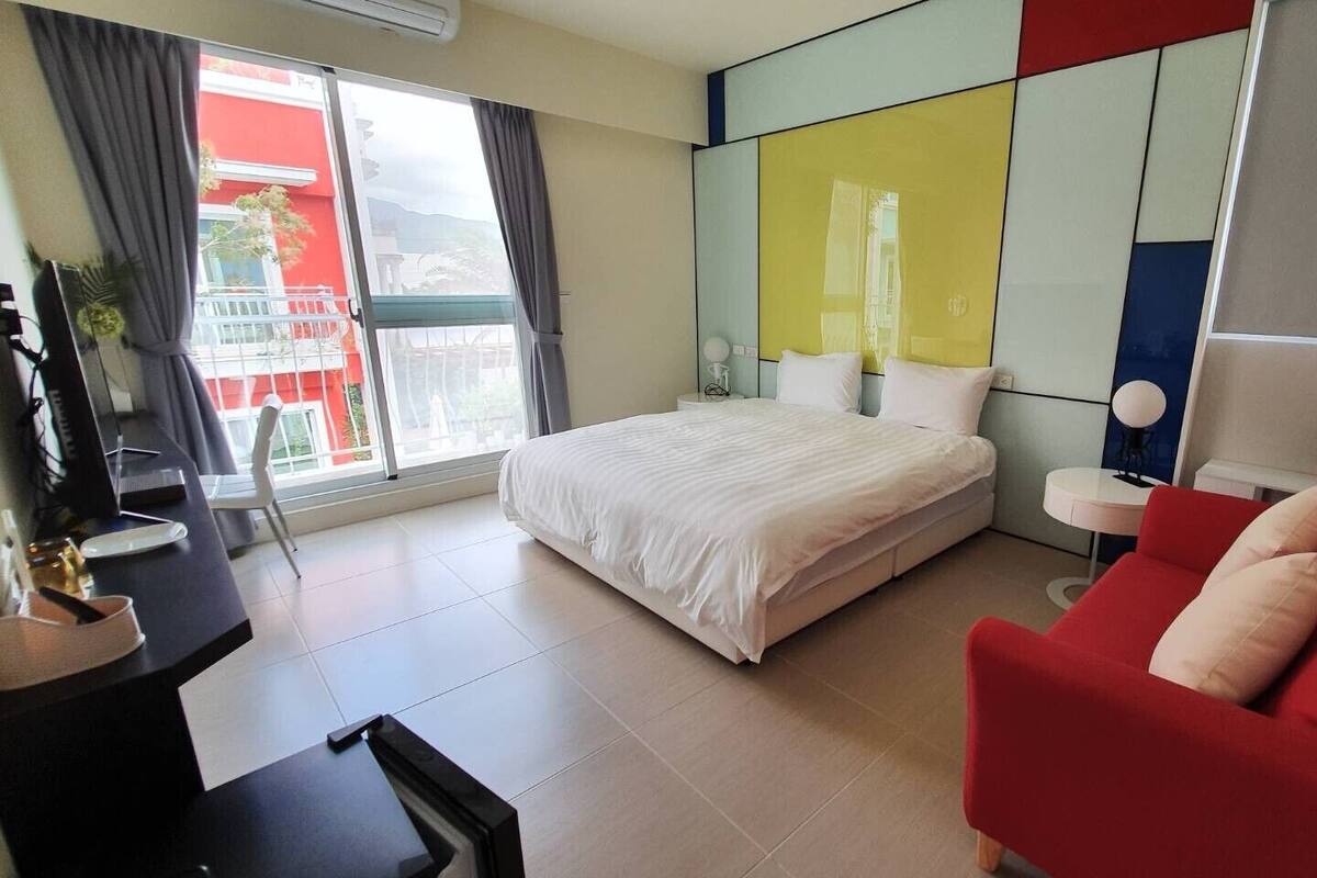 Quarto de hotel moderno com uma cama, um sofá vermelho, uma mesinha e uma janela com cortinas.
