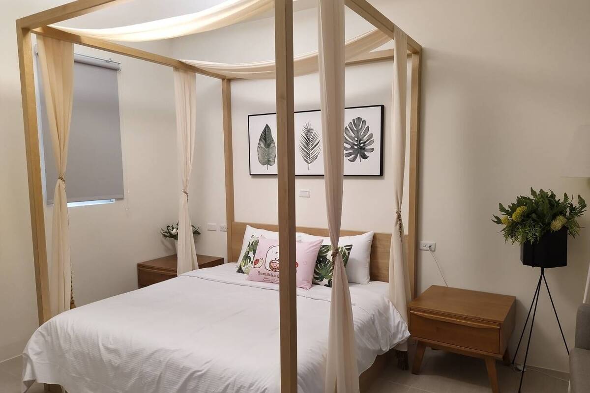 Quarto com cama de dossel, mesa de cabeceira, planta em vaso e impressões botânicas emolduradas na parede.
