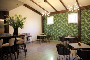 Restaurante com papel de parede tropical, vigas de madeira e um candelabro. Mesas e cadeiras dispostas para refeições.