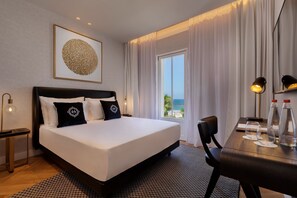 Quarto de hotel com uma cama grande, uma escrivaninha com dois copos e garrafas, uma mesa de cabeceira com abajur e uma janela ampla com cortinas.