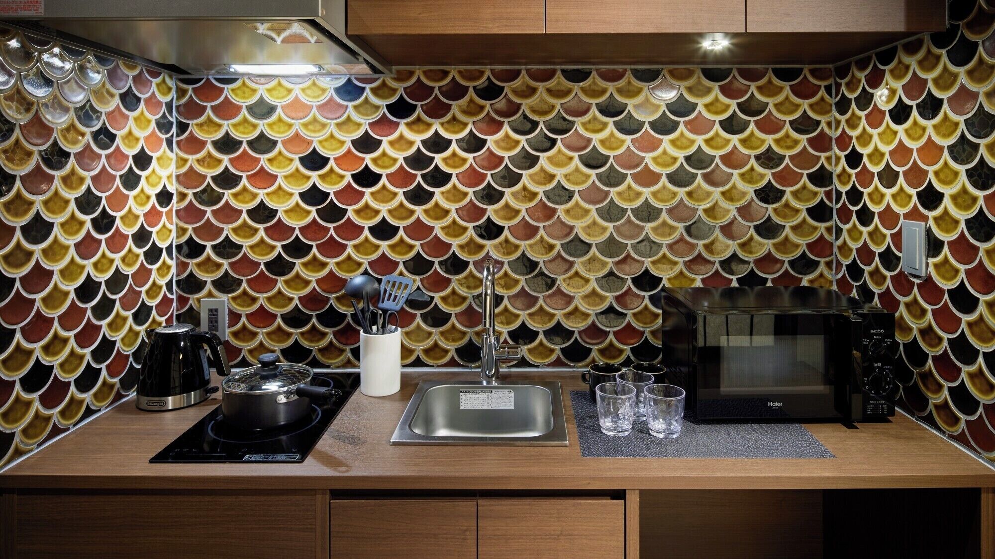 Cozinha moderna com painel de azulejos em formato de escamas de peixe, pia de aço inoxidável, fogão por indução preto, micro-ondas Haier e um conjunto de copos de vidro no balcão.