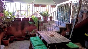 Uma área de pátio com mesa de madeira e assentos acolchoados verdes, rodeada por plantas em vasos e uma escada de metal.