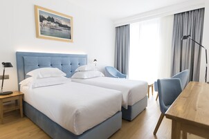 Quarto de hotel com duas camas, uma escrivaninha, uma cadeira, um abajur e uma janela com cortinas.