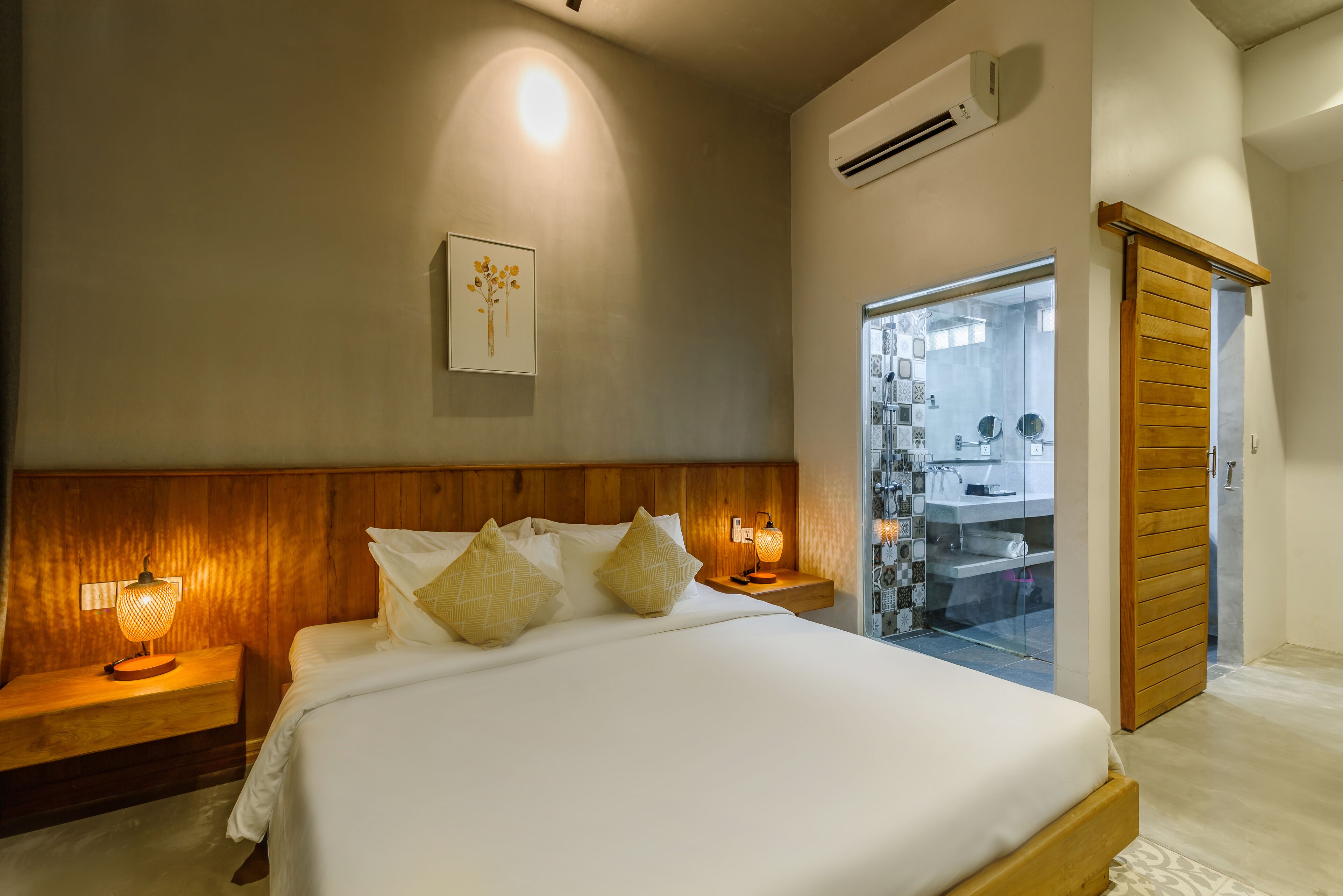 Quarto com cabeceira de madeira, mesa de cabeceira com abajur, cama com roupa de cama branca e almofadas estampadas, e box de chuveiro em vidro.