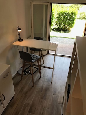 Espaço de escritório compacto com escrivaninha, cadeira e luminária. Uma janela oferece vista para o verde lá fora.