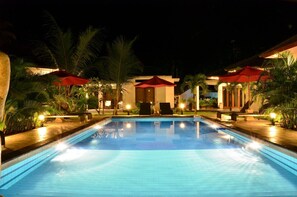 Uma área de piscina à noite, com uma piscina retangular, guarda-sóis vermelhos e palmeiras.