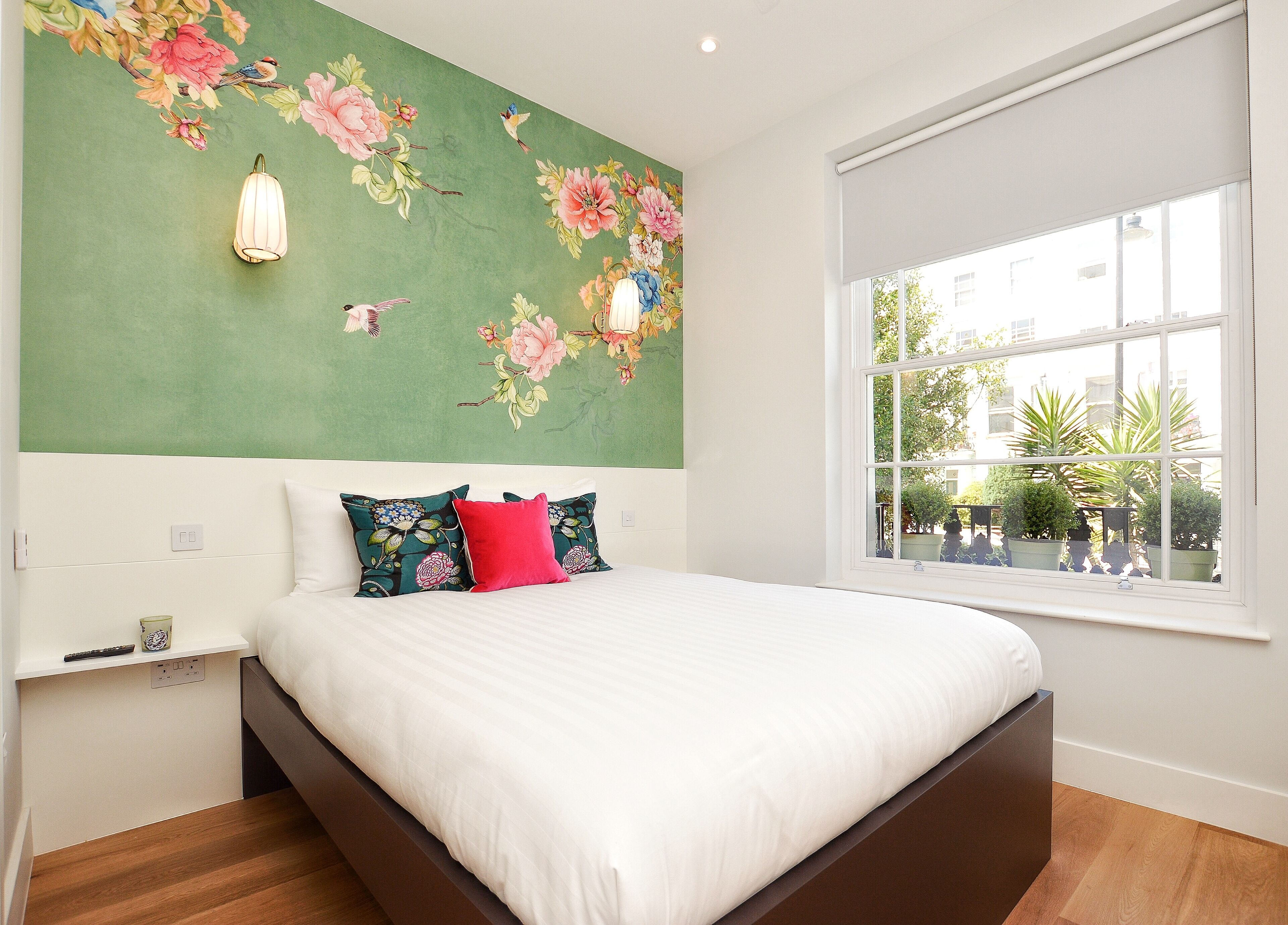 Quarto com parede verde, papel de parede floral, uma janela grande e uma cama com roupa de cama branca e almofadas coloridas.