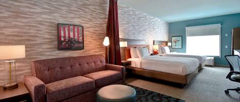 Quarto de hotel com cama, sofá, banquinho redondo, escrivaninha com luminária e janela com cortinas.