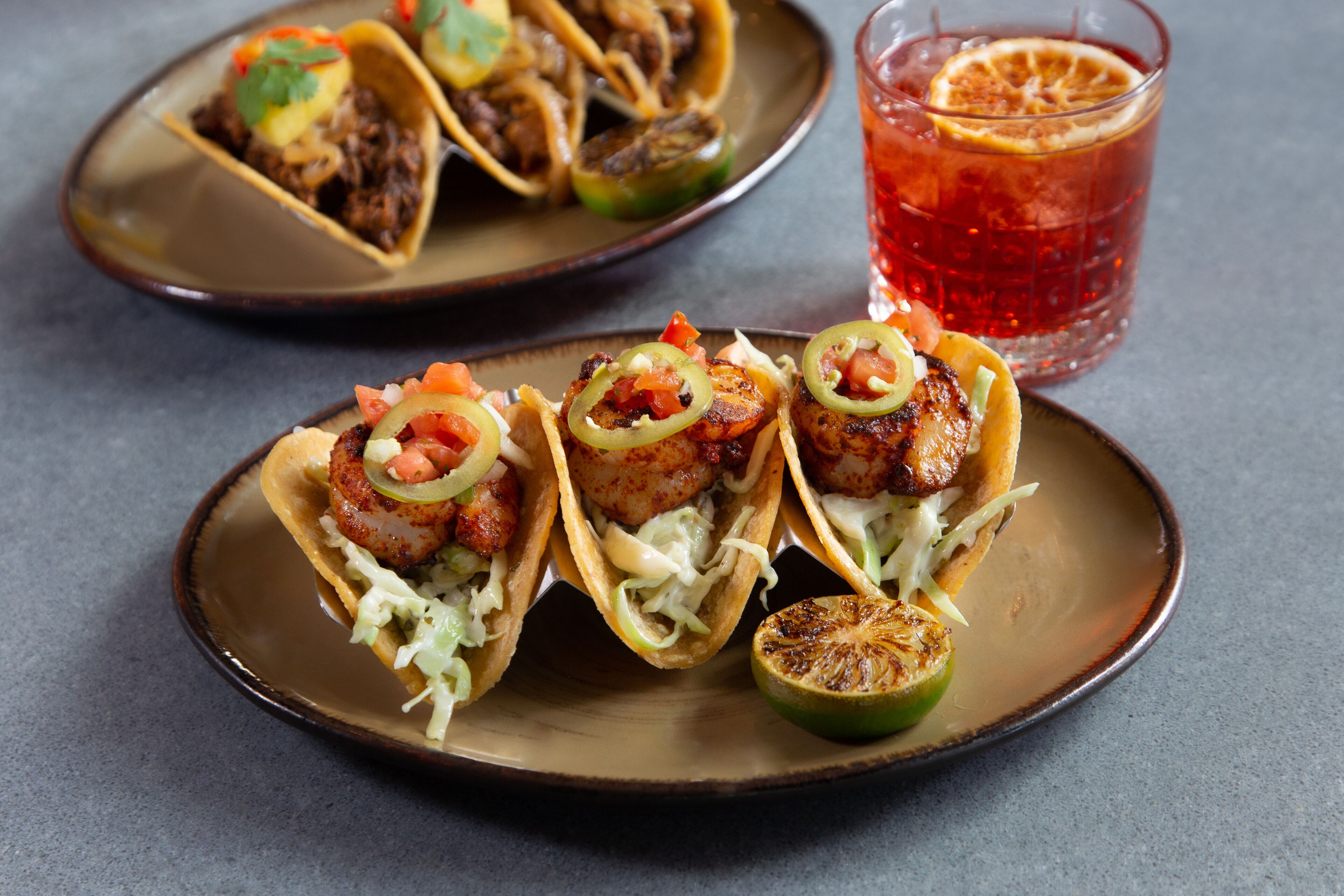 Três tacos com carne grelhada, cebolas e jalapeños em um prato, e um copo de bebida vermelha com uma fatia de laranja ao lado.
