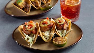 Três tacos com carne grelhada, cebolas e jalapeños em um prato, e um copo de bebida vermelha com uma fatia de laranja ao lado.