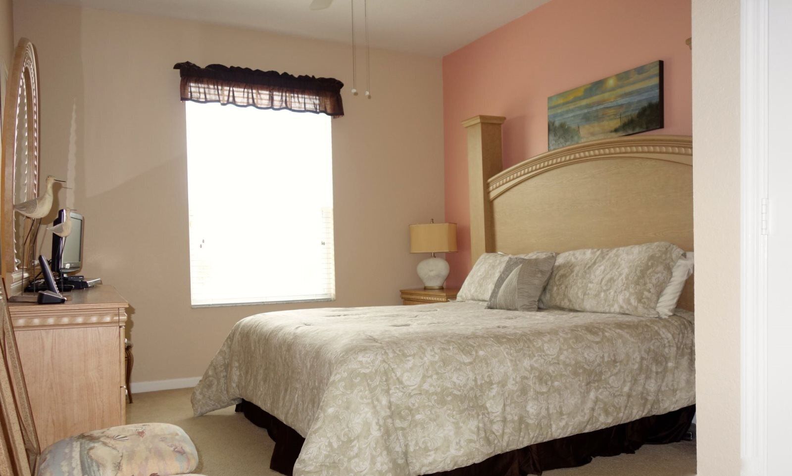 Um quarto com cama, penteadeira, luminária, pintura e janela com cortina.