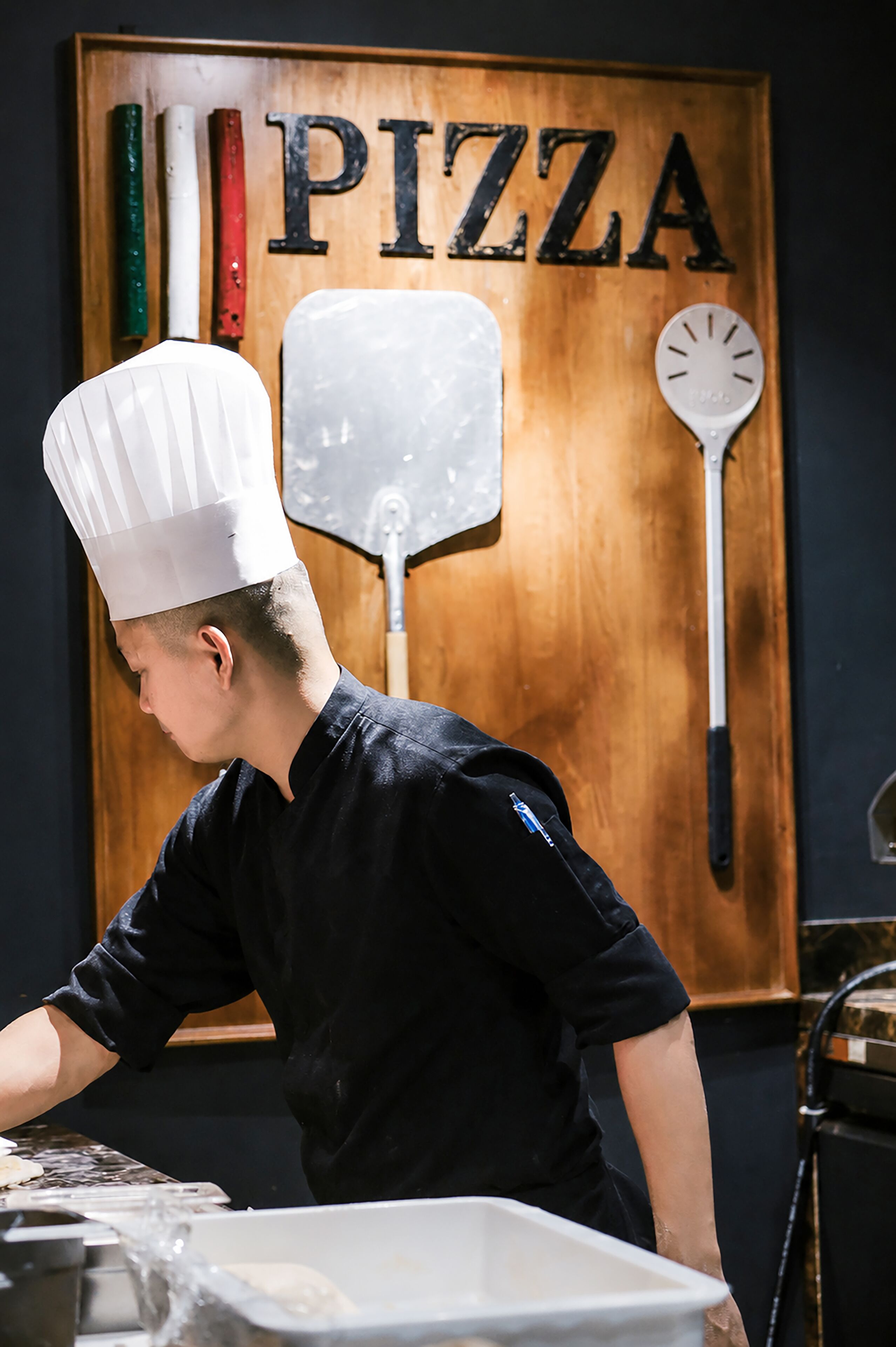 Um cozinheiro com uniforme preto e chapéu branco prepara comida em uma cozinha com parede de madeira e uma placa que diz 'PIZZA'.