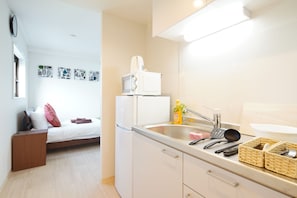 Cozinha compacta com micro-ondas, pia e diversos utensílios. Uma cama com travesseiro é visível ao fundo.