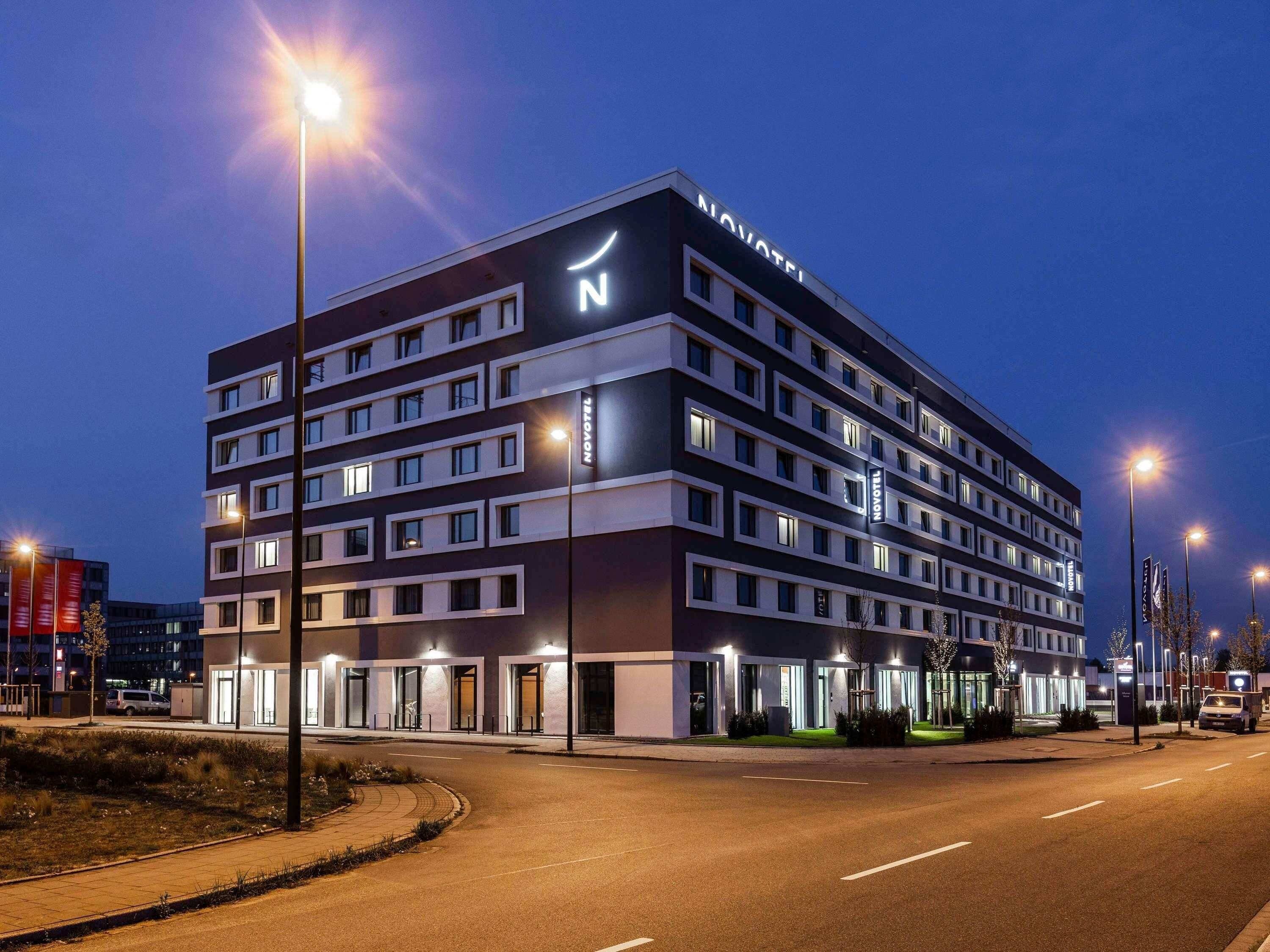 Um hotel de vários andares com design moderno, destacando uma placa grande com a inscrição 'NOVOTEL', várias janelas por andar e uma área de entrada bem iluminada.