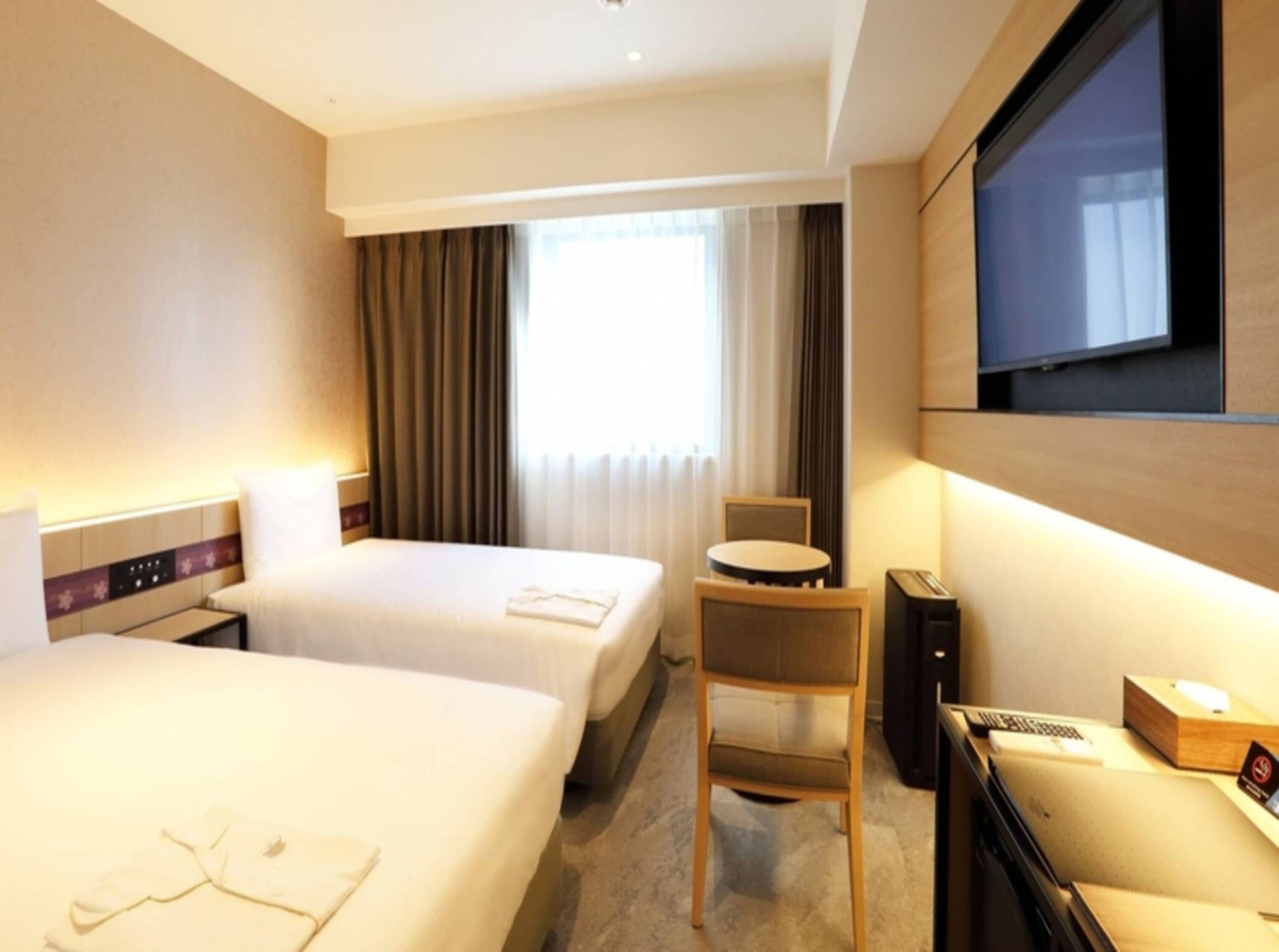 Quarto de hotel com duas camas, uma escrivaninha, uma cadeira, uma televisão e janela com cortinas.