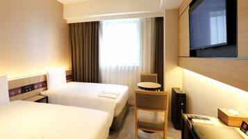 Quarto de hotel com duas camas, uma escrivaninha, uma cadeira, uma televisão e janela com cortinas.