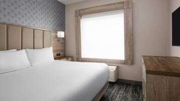 Quarto de hotel com uma cama grande, cabeceira de madeira, mesa de cabeceira com abajur, janela com persianas e cómoda de madeira.