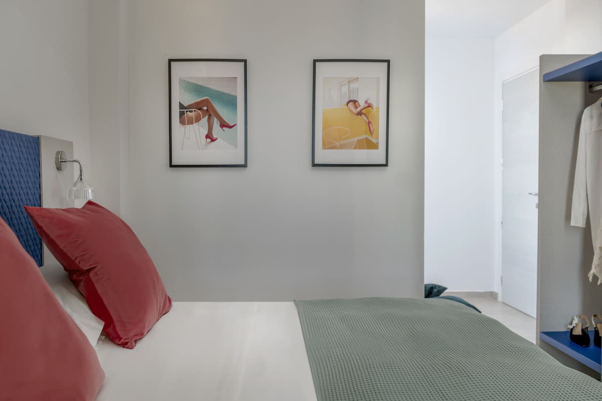 Quarto moderno com cama, almofadas vermelhas e verdes, cabeceira azul e dois quadros na parede.