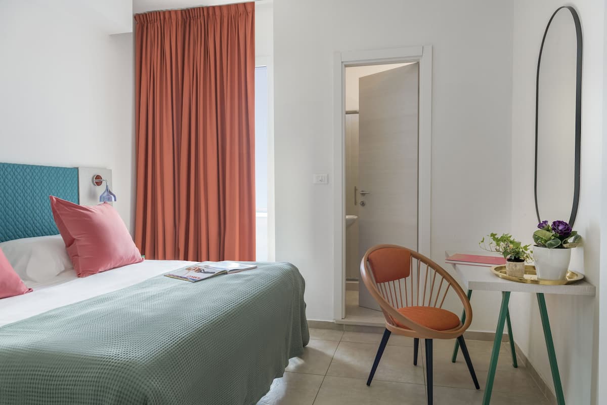 Quarto de hotel com cama, poltrona, escrivaninha e porta.
