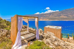Um gazebo de madeira com uma cama, com vista para uma área costeira com mar azul límpido e montanhas ao longe.