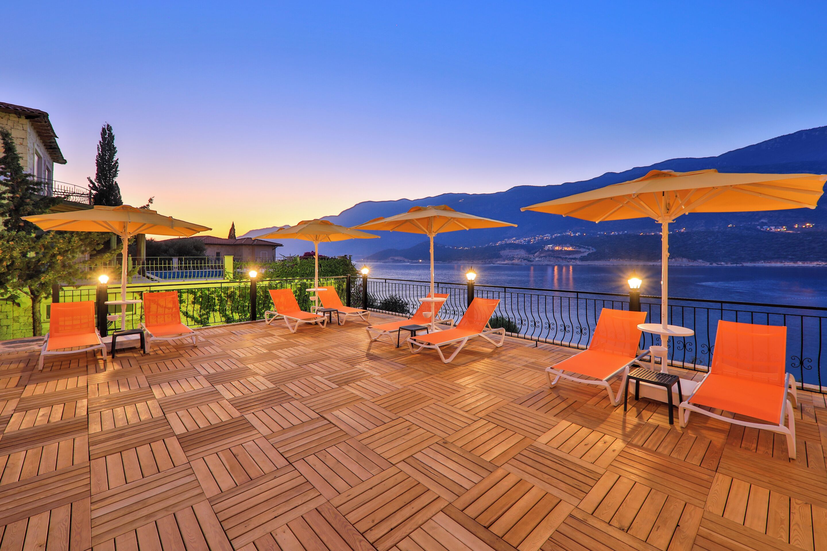Um deck de madeira com cadeiras de lounge laranja e guarda-sóis, com vista para um lago e montanhas ao fundo.