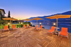 Um deck de madeira com cadeiras de lounge laranja e guarda-sóis, com vista para um lago e montanhas ao fundo.