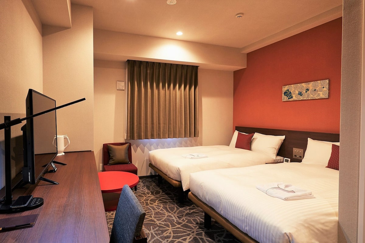 Quarto de hotel com duas camas, uma escrivaninha com televisão, uma cadeira vermelha e um tapete estampado.