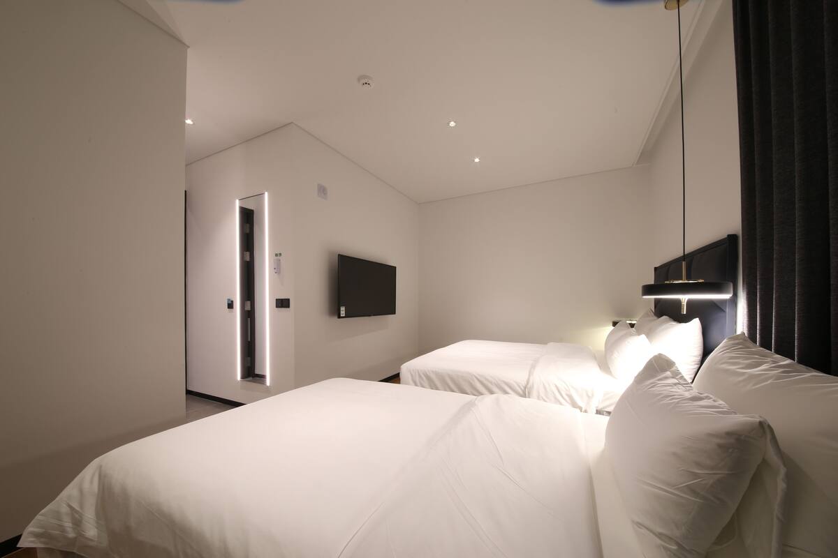 Quarto de hotel moderno com duas camas, uma televisão de tela plana na parede e uma porta que dá para outro cômodo.