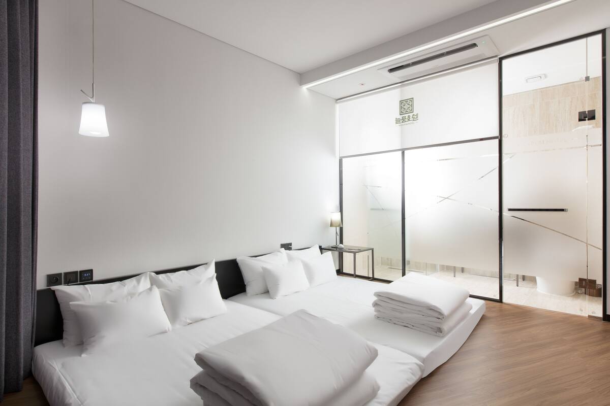 Quarto de hotel moderno com uma cama grande, mesa de cabeceira, espelho e porta de vidro deslizante para um banheiro.