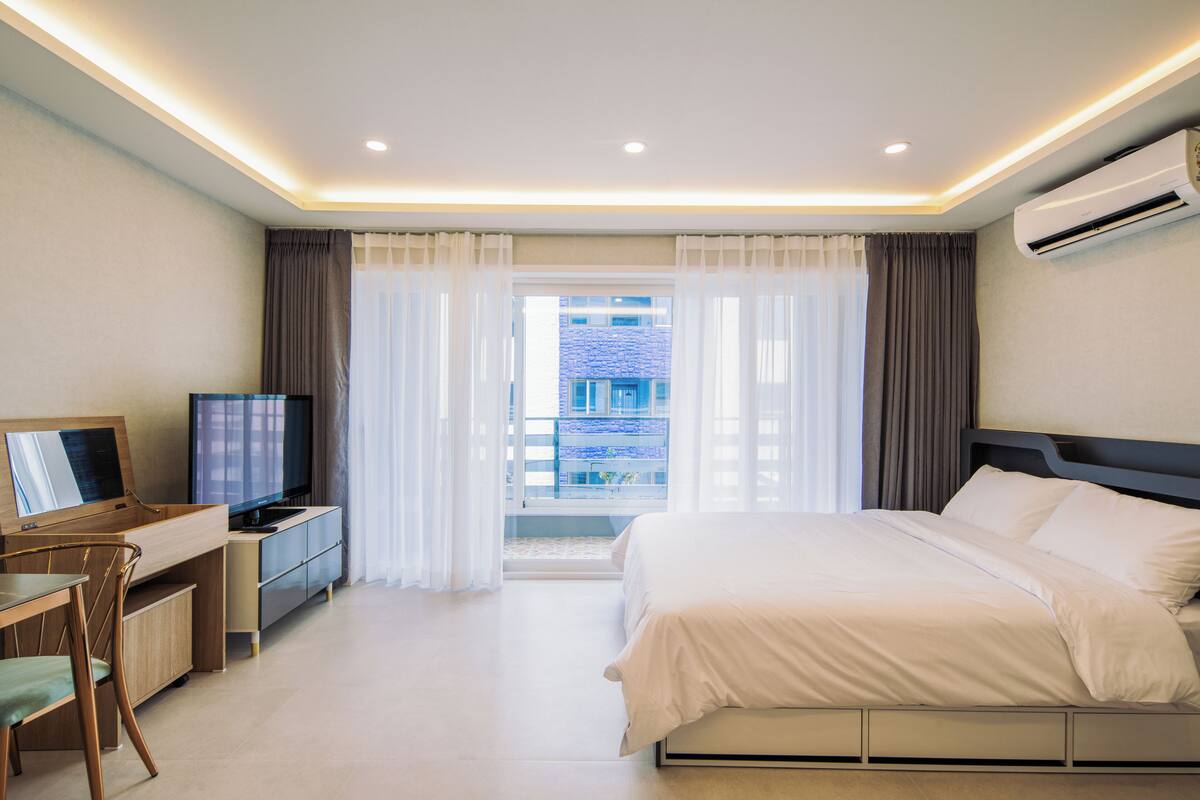 Quarto de hotel com cama, escrivaninha com televisão, janela com cortinas e ar condicionado.