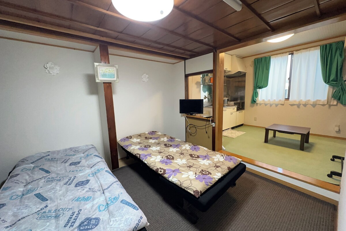 Quarto pequeno e funcional com cama, escrivaninha, cadeira e um banco.