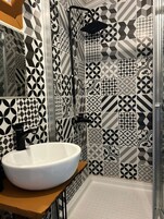 Casa de banho com lavatório branco, torneira preta e chuveiro com mangueira preta. As paredes são revestidas com padrões geométricos em preto e branco.