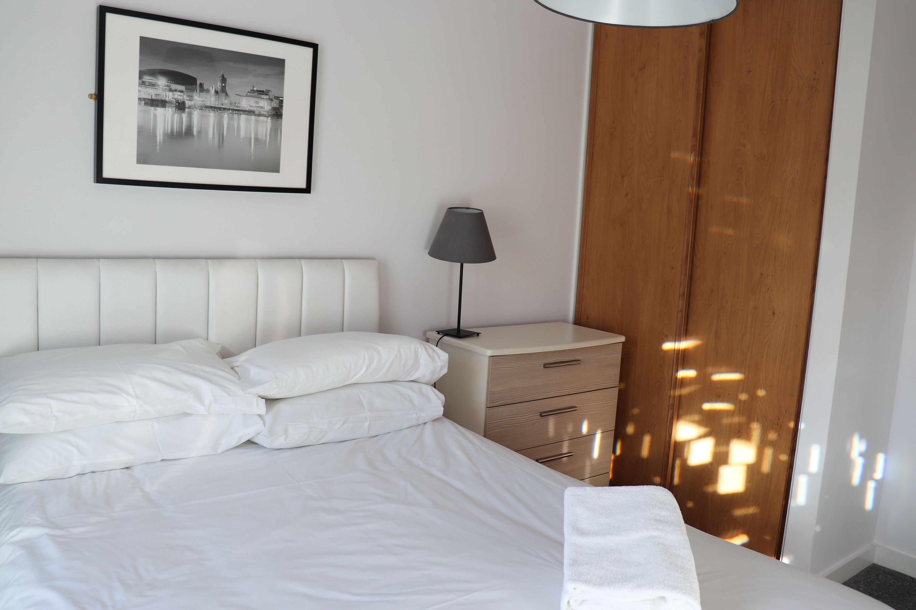 Cama bem arrumada com lençóis brancos, abajur e quadro na parede.
