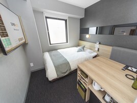 Quarto de hotel com cama, escrivaninha, pequeno espaço de cozinha e janela.