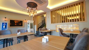 Interior moderno de restaurante com mesas de madeira, cadeiras estofadas cinzas, um candelabro e uma escadaria para um andar superior.