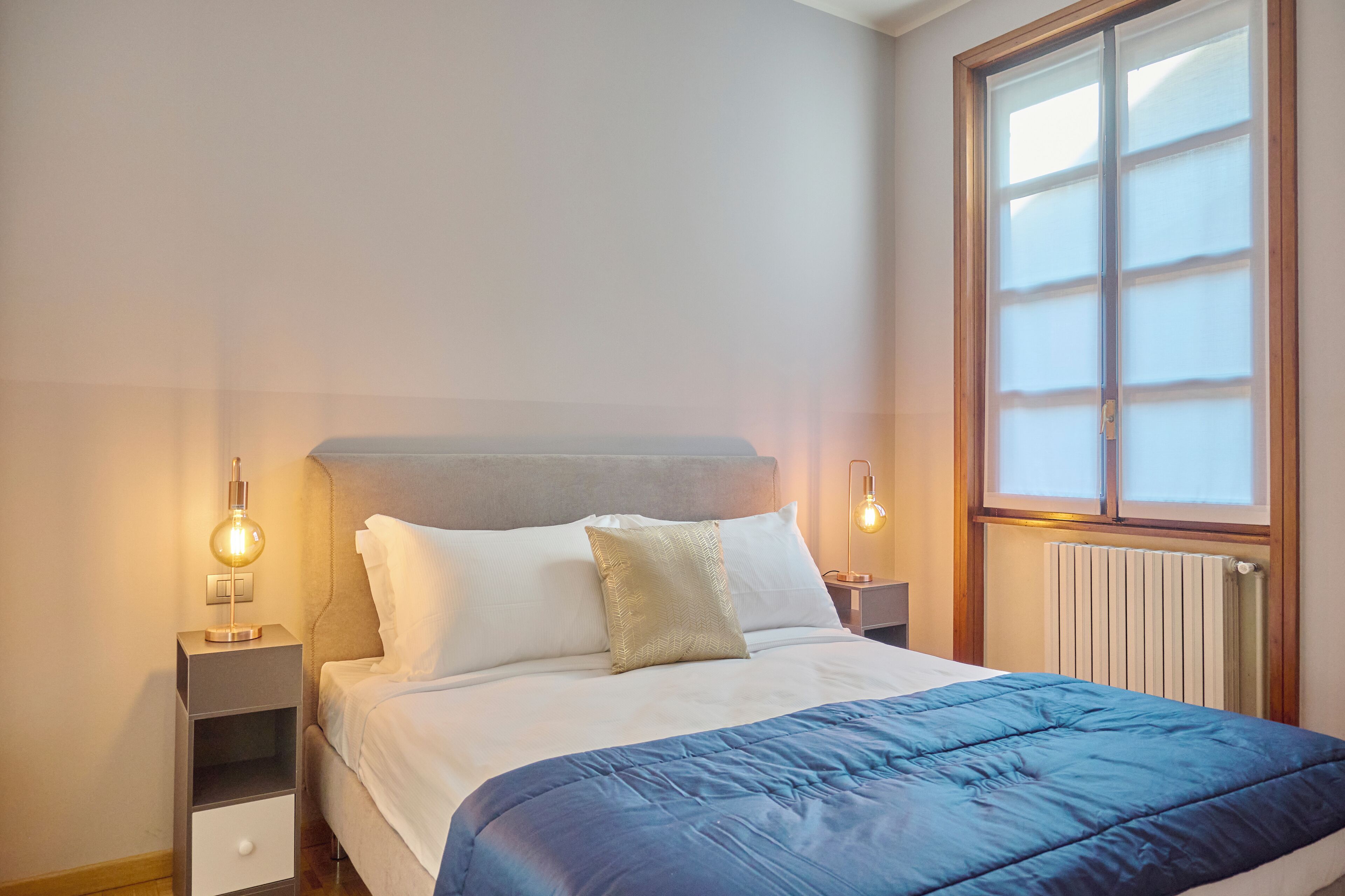 Uma cama bem arrumada com um cobertor azul, duas luminárias de cabeceira e uma janela com persianas.
