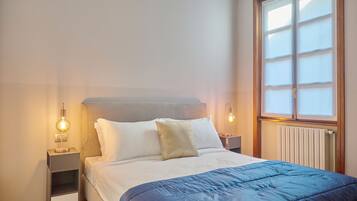 Uma cama bem arrumada com um cobertor azul, duas luminárias de cabeceira e uma janela com persianas.