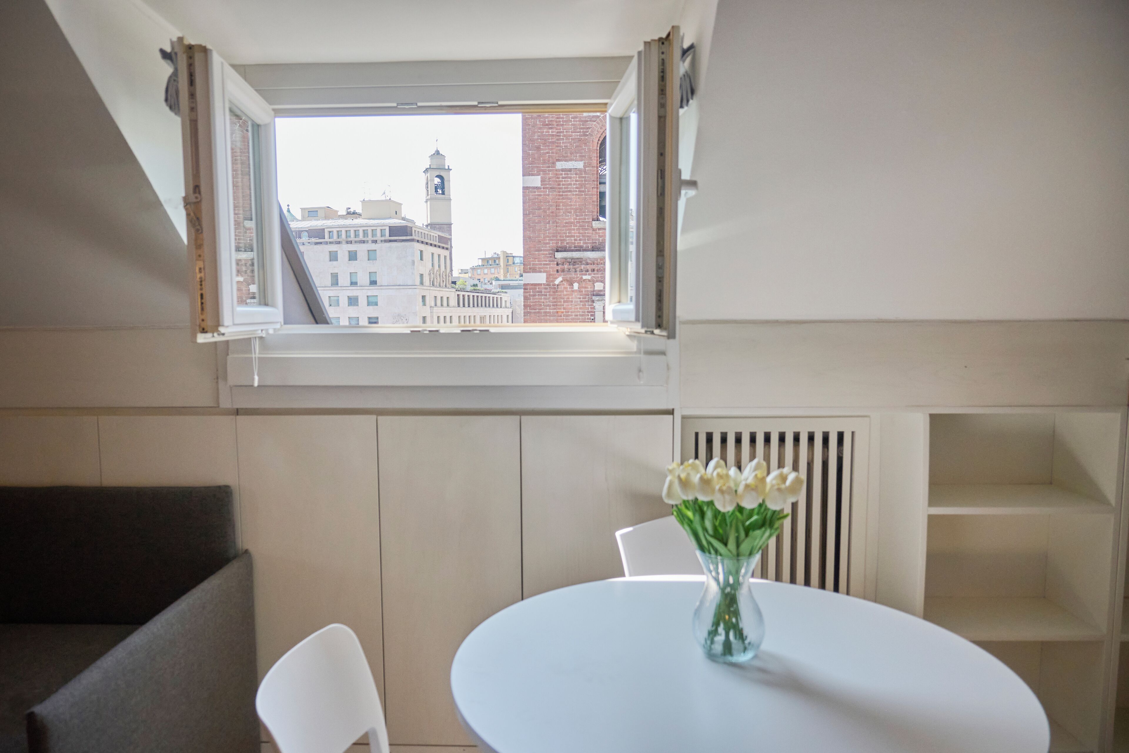 Um quarto com uma mesa branca, um vaso com flores, uma cadeira branca, um sofá cinzento e uma janela com vista para edifícios.
