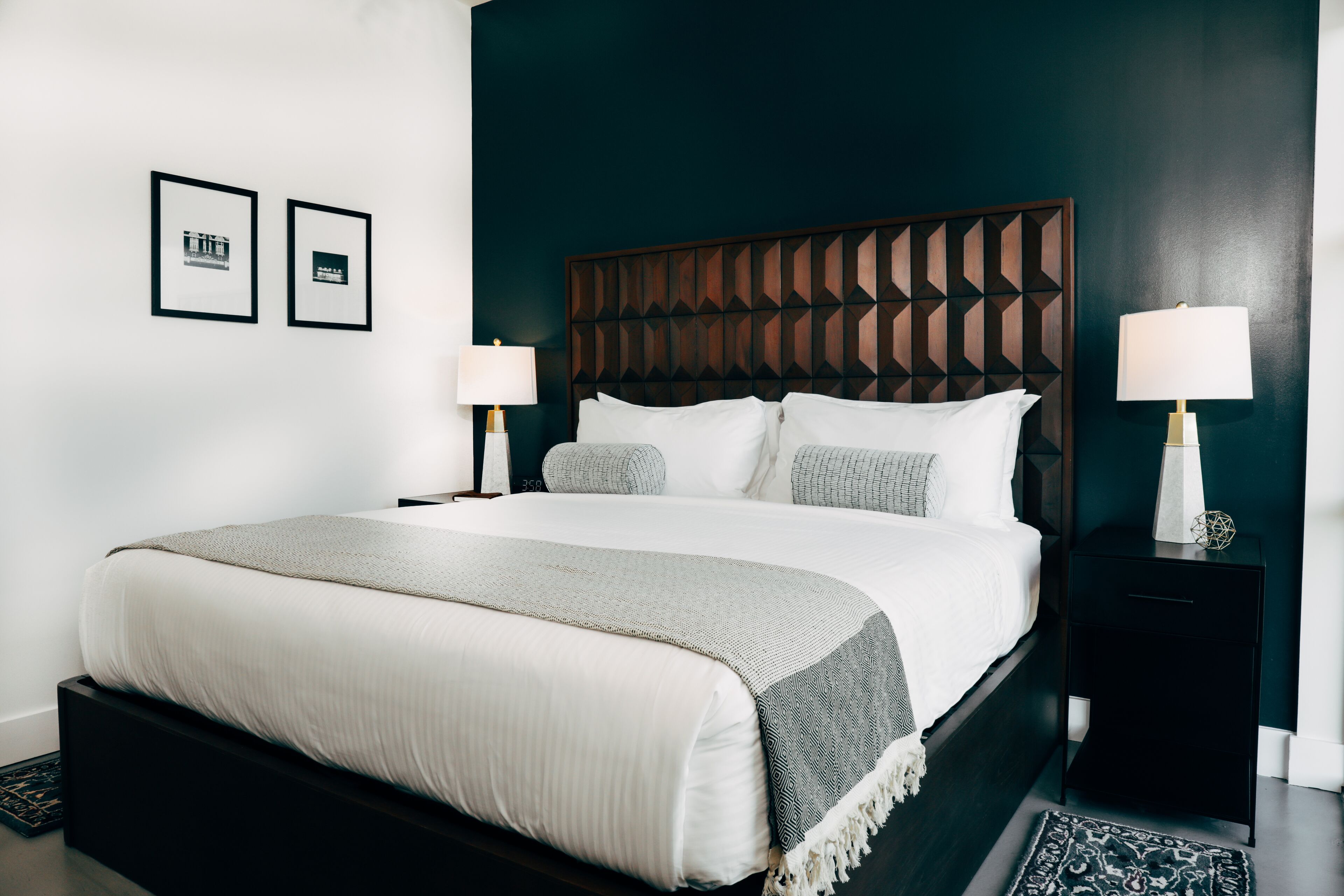 Cama bem arrumada com cabeceira de madeira, duas mesas de cabeceira com luminárias e quadros nas paredes.