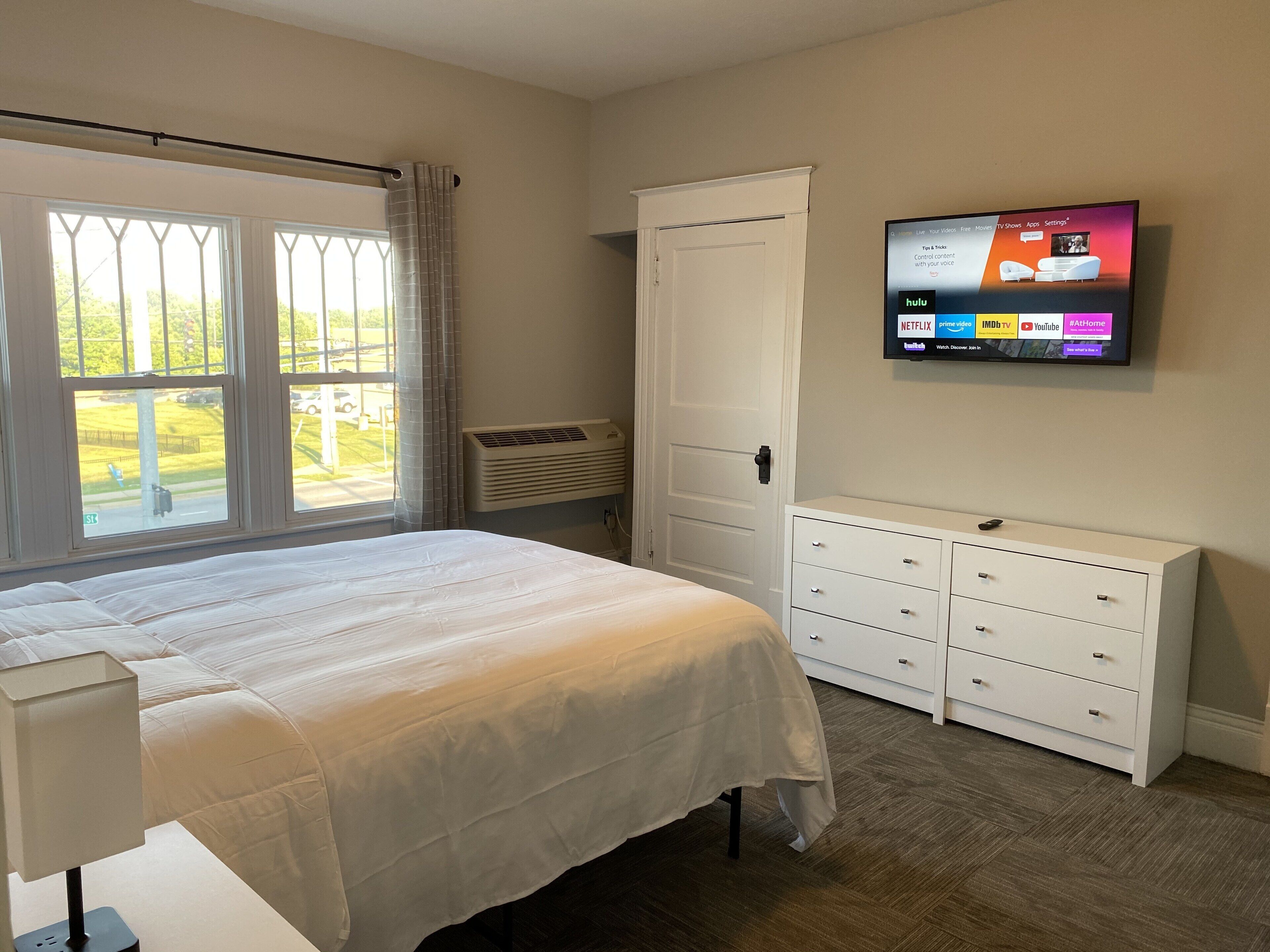 Quarto com cama, cômoda, TV na parede e janela com cortinas.